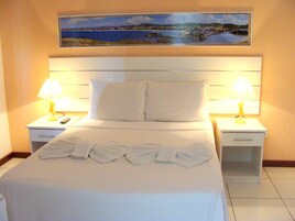 Quarto de hotel com cama bem arrumada, duas mesas de cabeceira com abajures, um quadro sobre a cama e vista para a paisagem costeira.