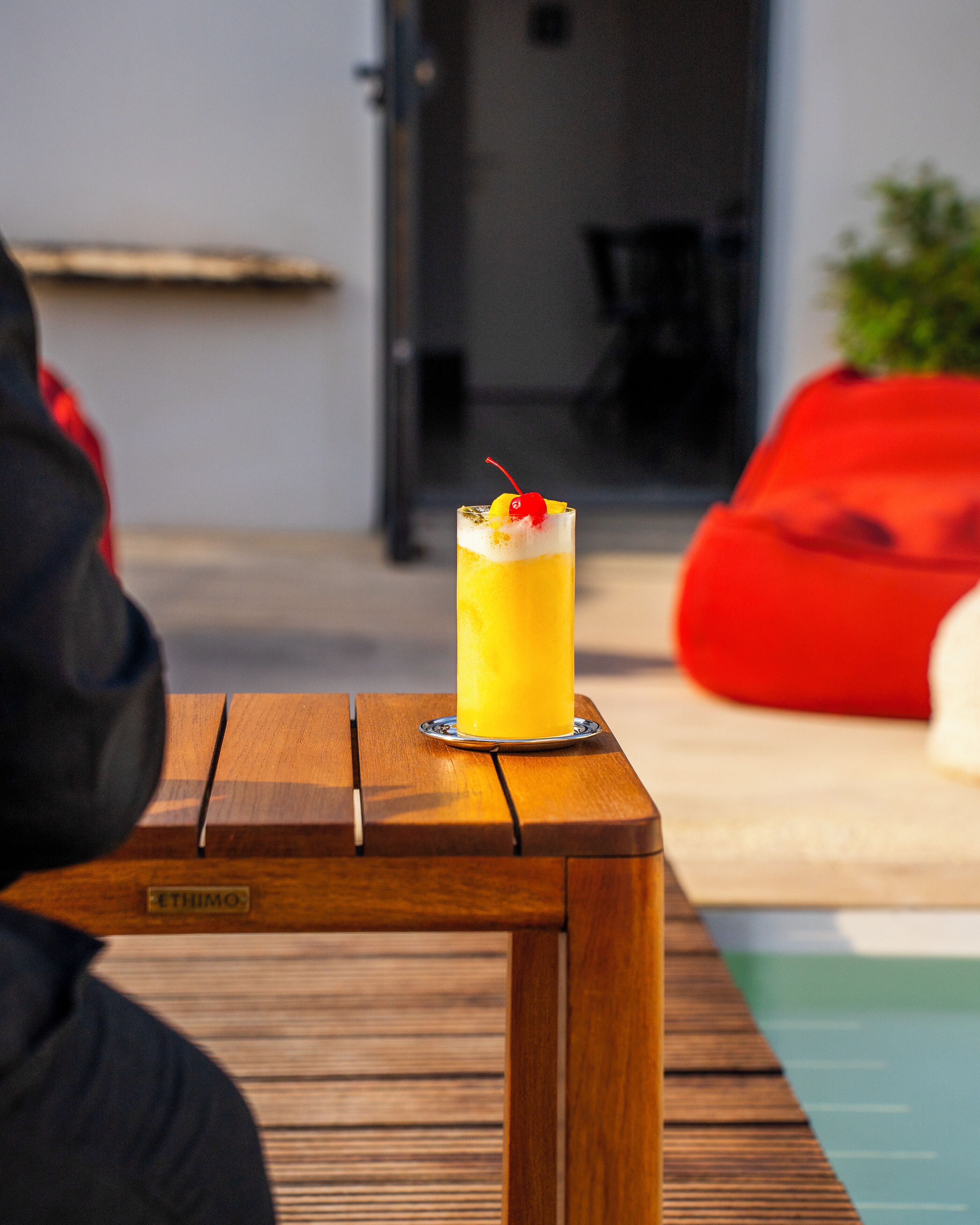 Uma mesa de madeira com um cocktail, um bean bag vermelho e uma piscina ao fundo.
