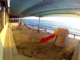 Uma praia com um parquinho, incluindo um escorregador e estruturas de escalada, sob uma área sombreada.