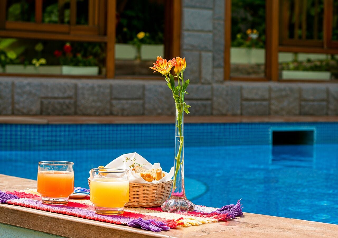 Disposição para café da manhã junto à piscina, com bebidas, pães e um vaso com flores.