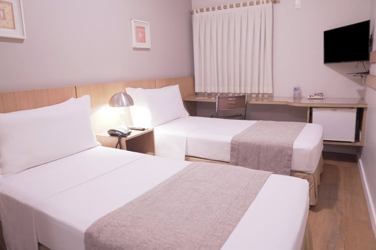 Quarto de hotel com duas camas, uma escrivaninha, uma cadeira, uma televisão e um frigorífico pequeno.