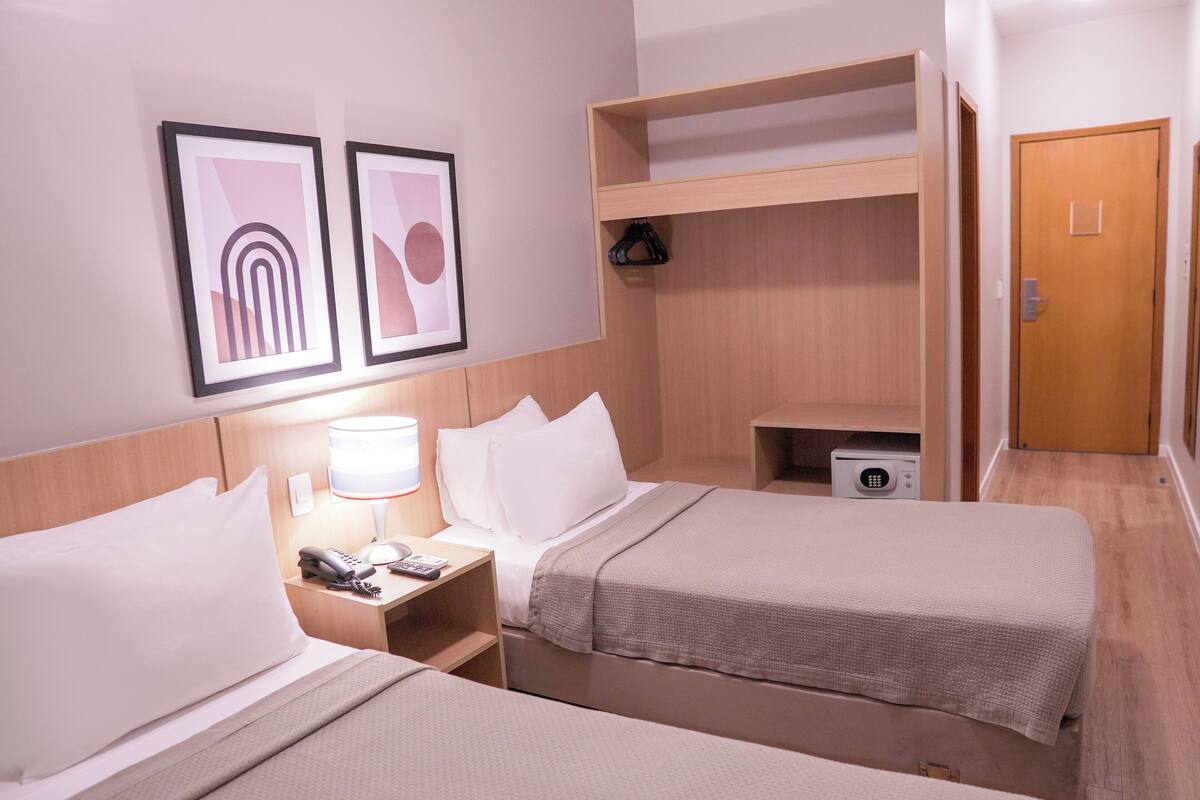 Quarto de hotel com duas camas, uma mesa de cabeceira com luminária, telefone e um cofre pequeno. Há quadros nas paredes e uma estante de madeira com um porta-casacos.