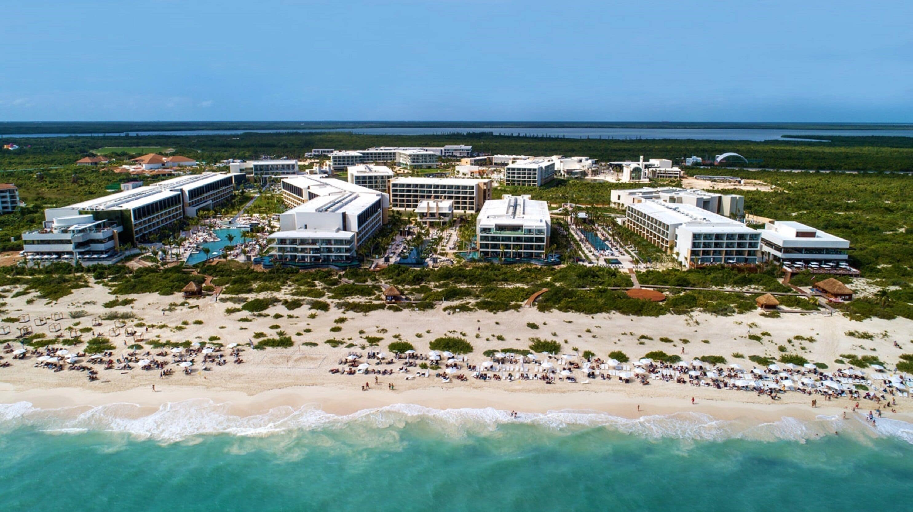 Vista aérea de um hotel à beira-mar, com vários edifícios, uma praia de areia e uma visão clara do oceano.
