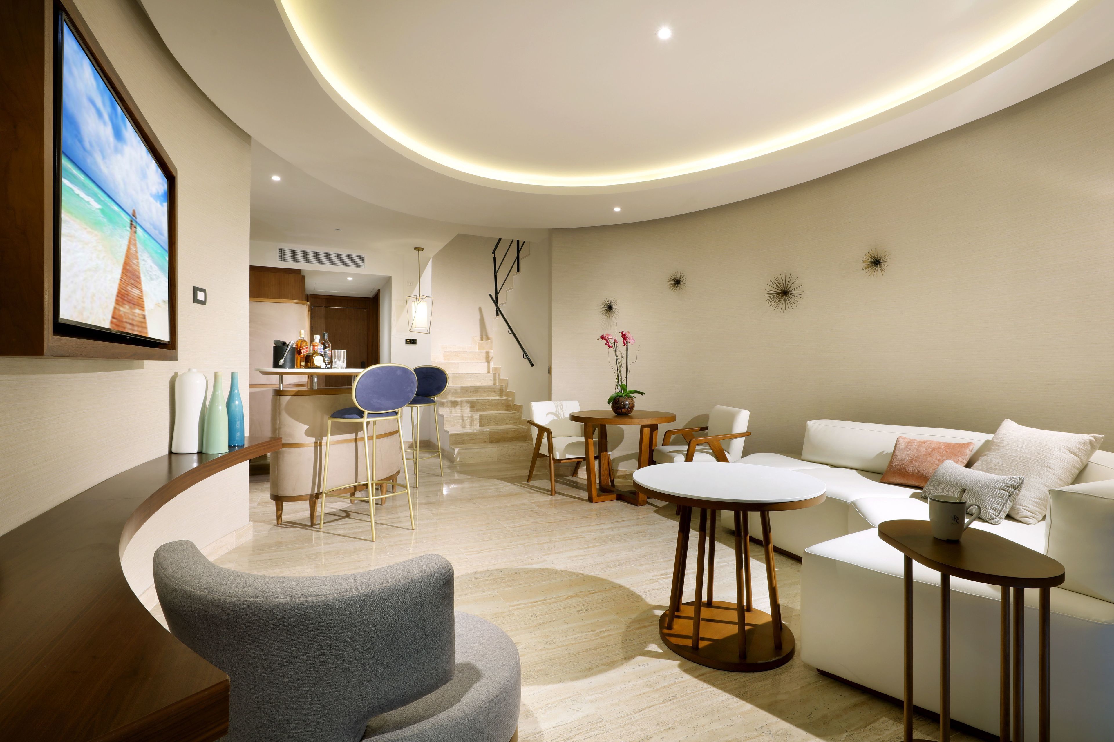 Uma sala de estar moderna com um balcão de recepção curvo em madeira, uma poltrona cinza, um sofá branco e uma pequena mesa redonda com uma planta.