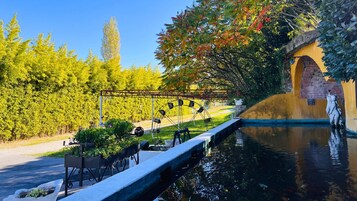 Um canal com uma ponte, uma pérgula com plantas e uma estátua.