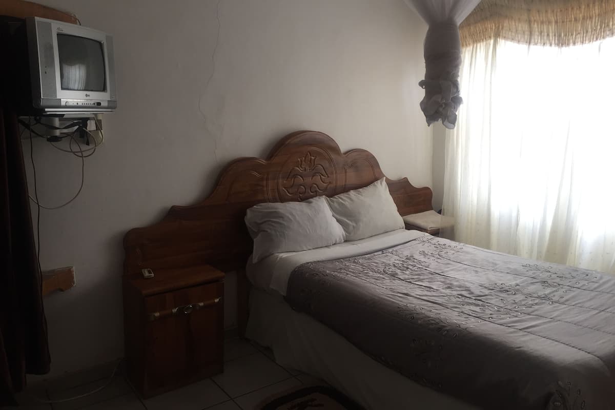 Quarto com cama de madeira, mesa de cabeceira, televisão fixada na parede e janela com cortinas leves.