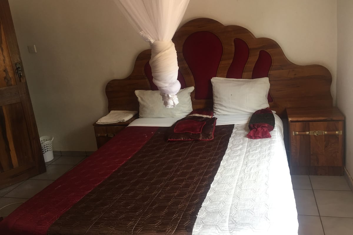 Quarto com cabeceira de madeira, cama com roupa de cama branca e vermelha, um tapete corrediço vermelho e uma mesinha de cabeceira de madeira com uma gaveta.