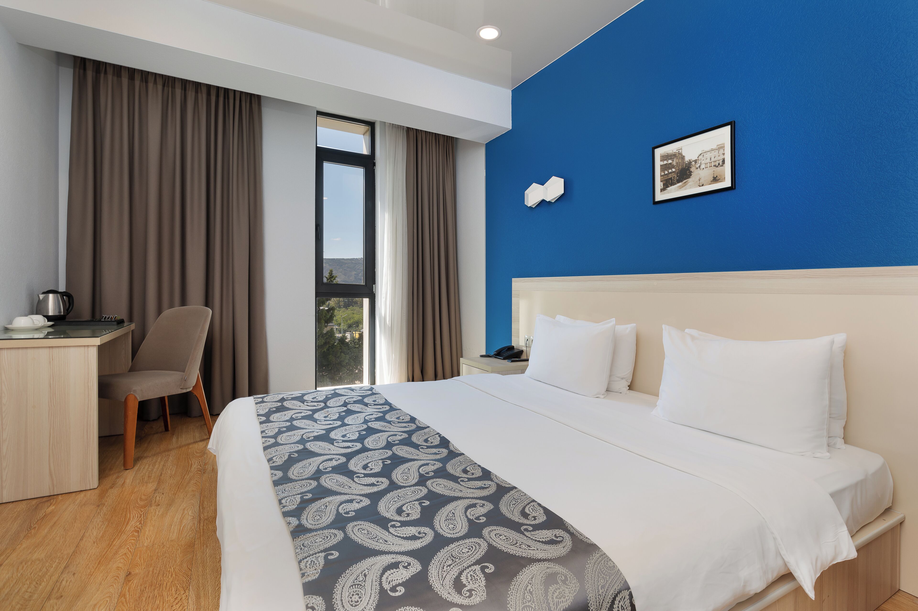 Quarto de hotel com cama, escrivaninha, cadeira e janela com cortinas.