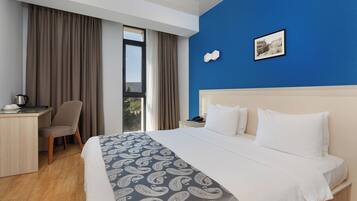 Quarto de hotel com cama, escrivaninha, cadeira e janela com cortinas.