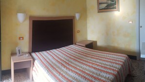 Quarto de hotel com uma cama grande, duas mesas de cabeceira, um criado-mudo, um quadro na parede e uma porta.