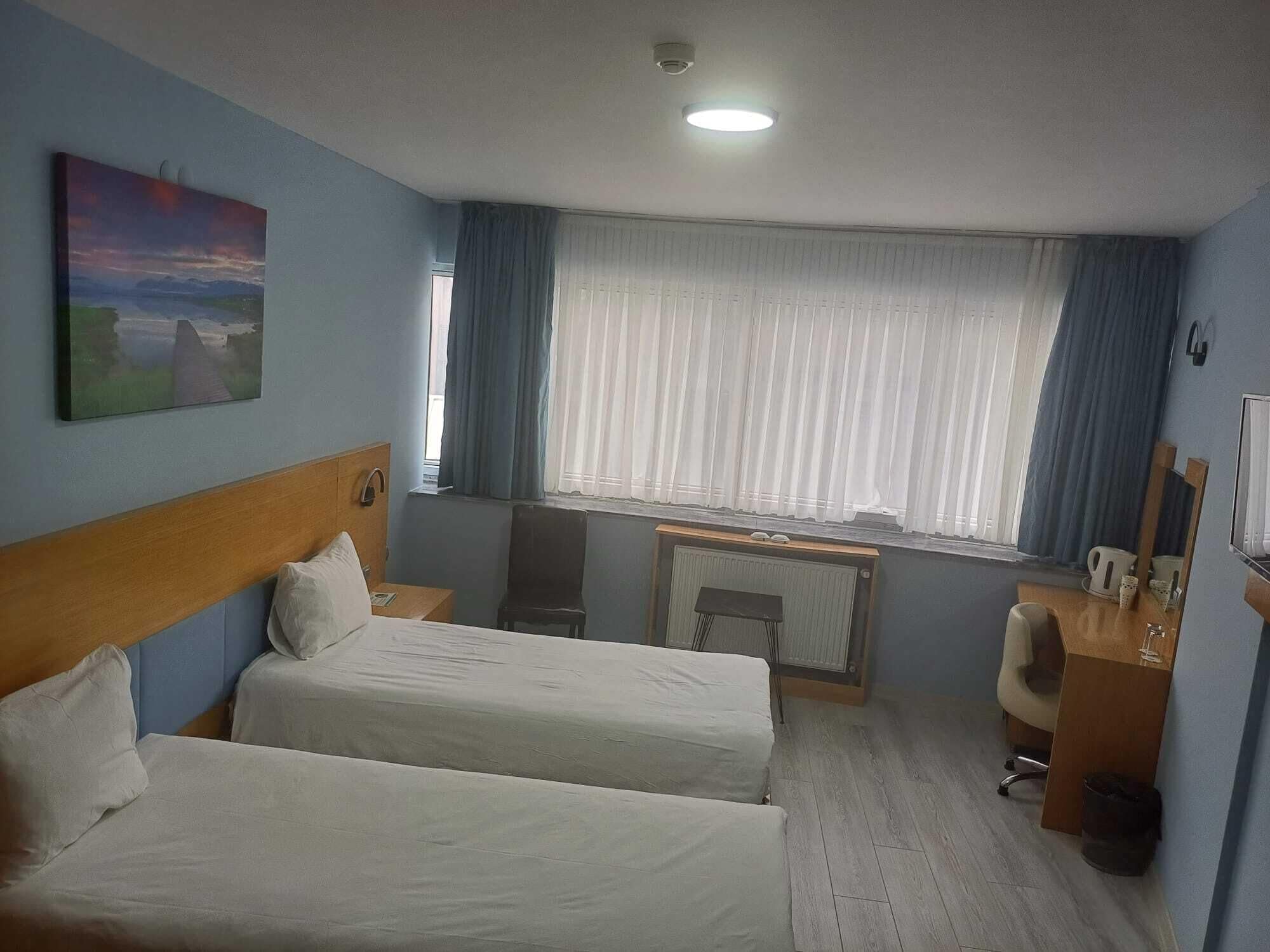Quarto de hotel com duas camas, uma escrivaninha, uma cadeira, um radiador e uma janela com cortinas.