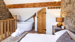 Quarto com cama grande, um banco de madeira, banheira branca e parede de pedra.