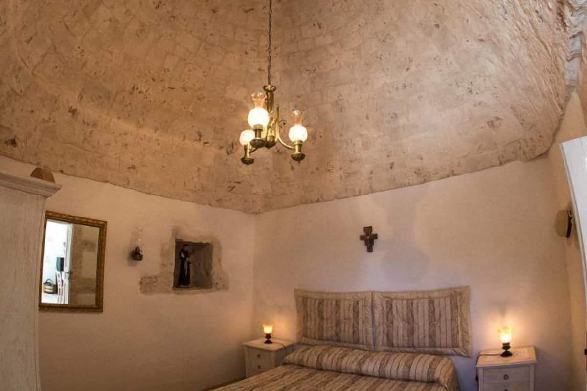 Quarto com teto abobadado, cama com colcha listrada, luminárias de cabeceira e uma pequena mesa de apoio.