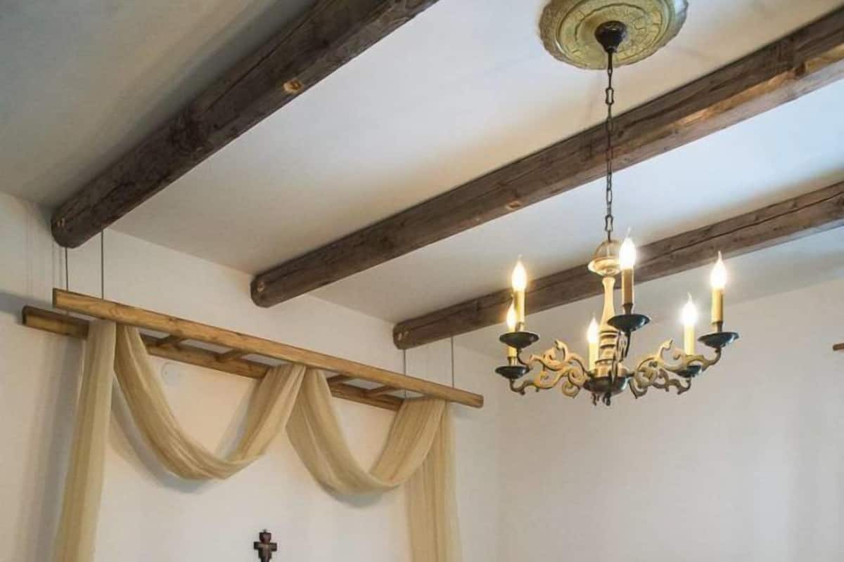 Quarto com cama, luminária de cristal, cortinas com pregas, cabeceira, mesa de cabeceira com abajur e relógio fixado na parede.