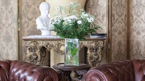 Um quarto com um sofá de couro marrom, um vaso de vidro com flores brancas e um busto em uma mesa.