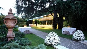 Uma trilha no jardim leva a uma área de refeições coberta, com mesas e cadeiras, cercada por vegetação e um grande vaso.