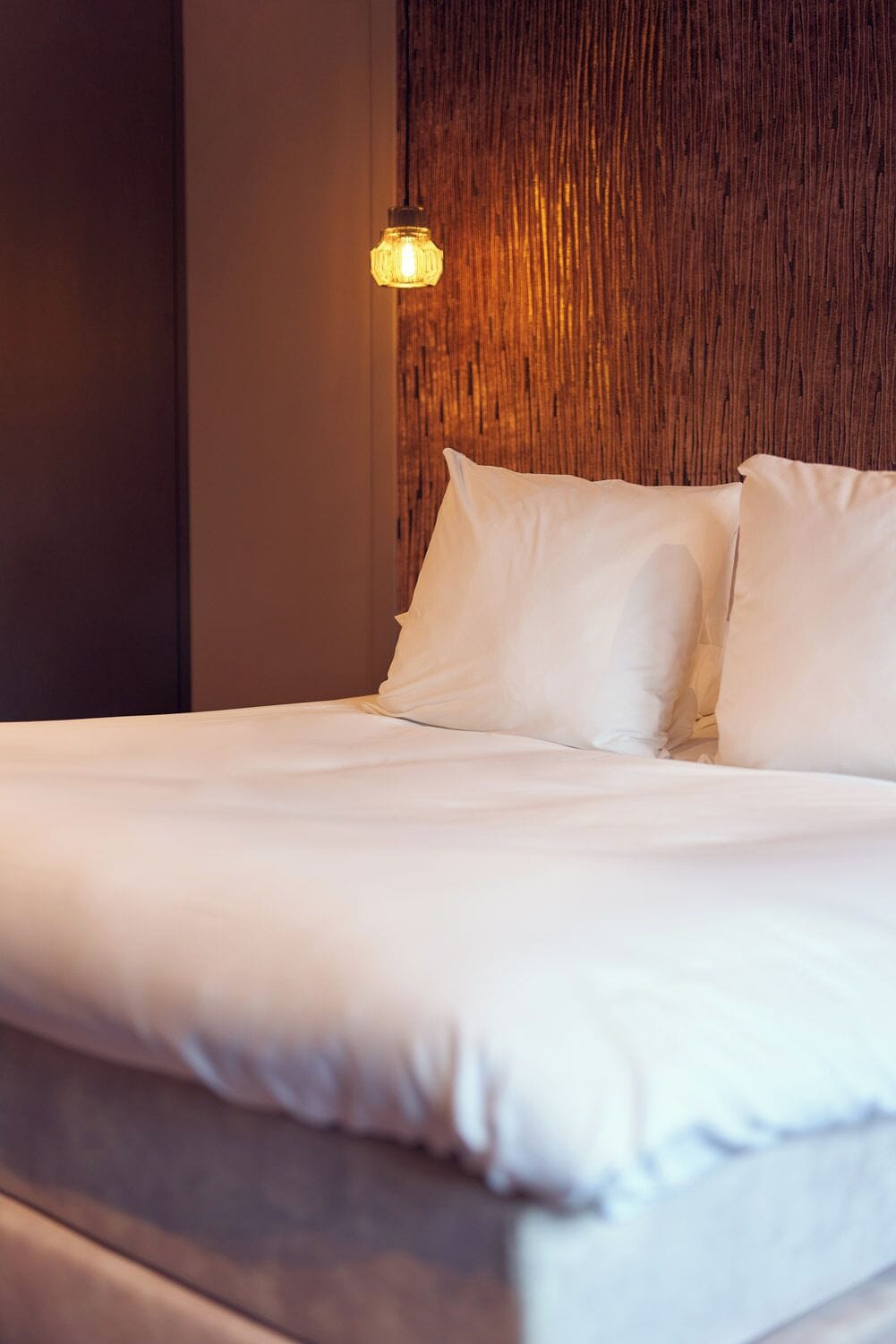 Cama bem arrumada com lençóis e almofadas brancos, cabeceira de madeira e luminária fixada na parede.