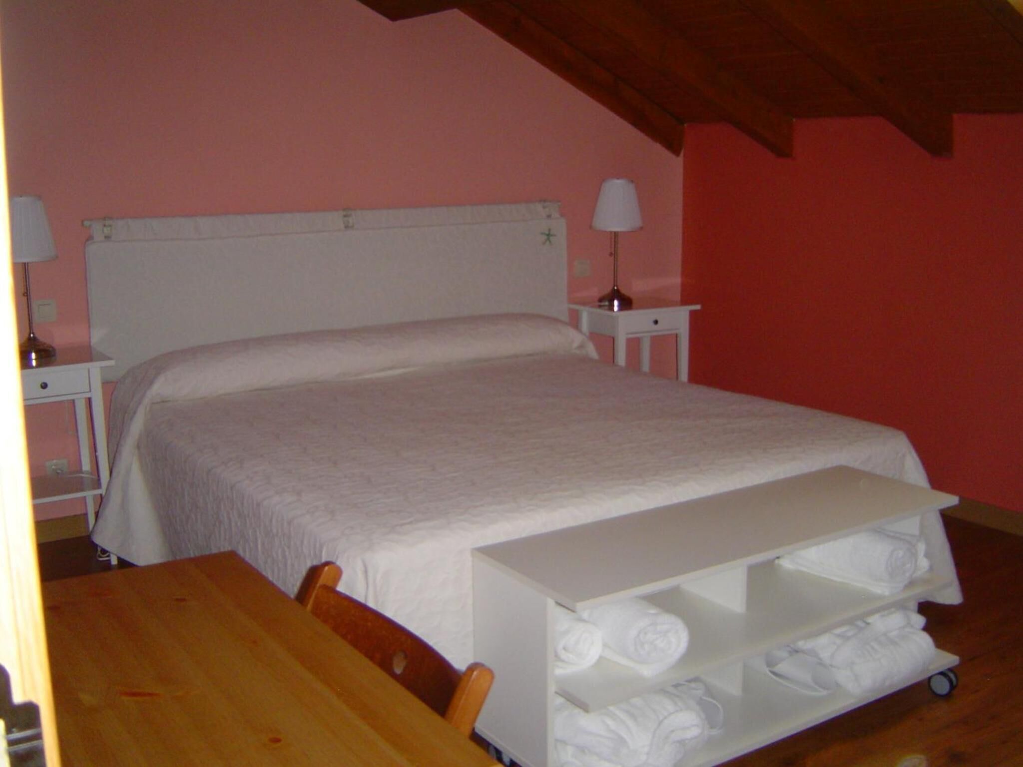 Quarto com cama, mesa de cabeceira, luminária, cadeira de madeira e prateleira com toalhas.