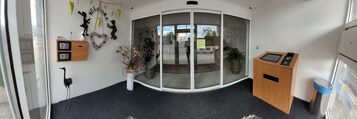Uma área de recepção de hotel com porta de vidro, balcão de madeira, lixeira e um quadro de informações fixado na parede.
