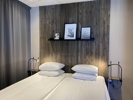 Quarto de hotel com cabeceira de madeira, duas camas com roupa de cama branca, uma estante com quadros emoldurados e abajures.