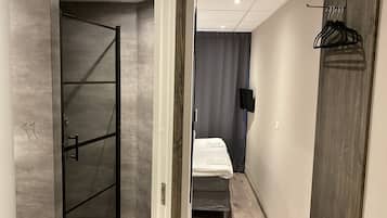 Um corredor estreito com uma porta de correr que dá para um quarto. O quarto possui uma cama, uma mesa de cabeceira e uma TV fixada na parede.