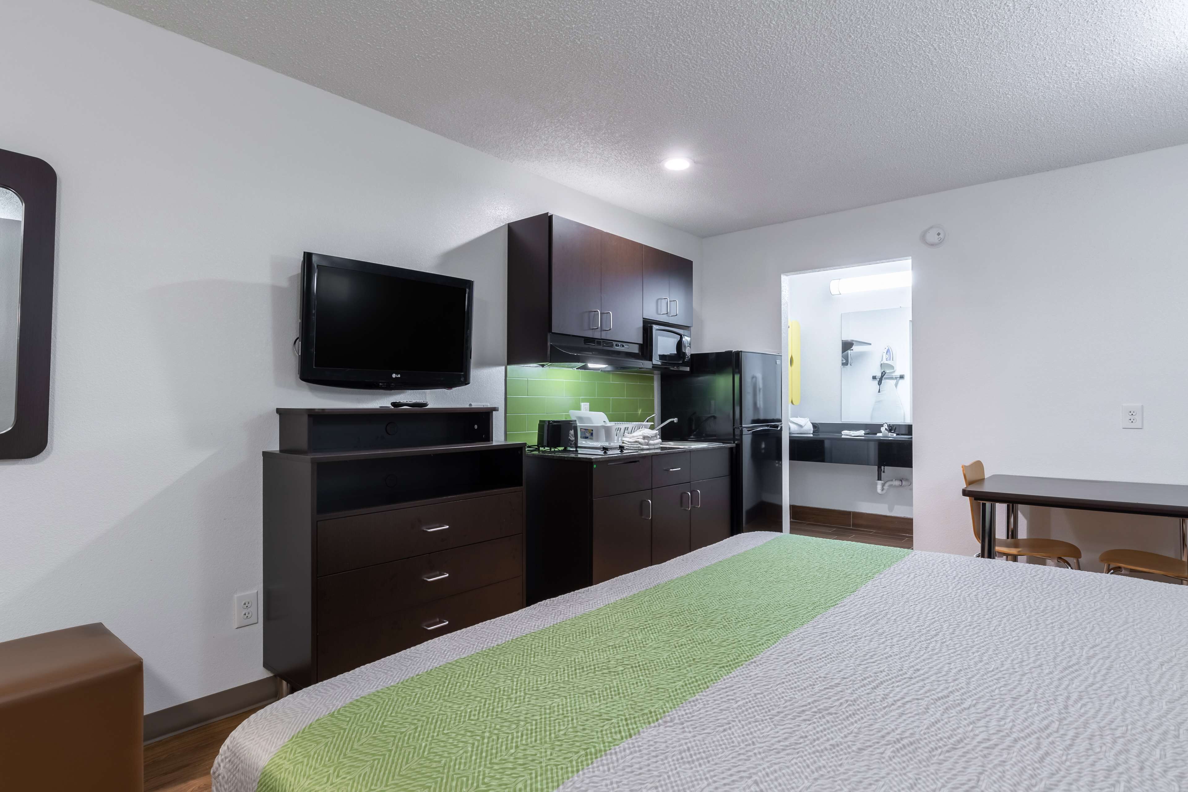 Quarto de hotel pequeno com cama, televisão, cozinha compacta e banheiro visível por uma porta aberta.