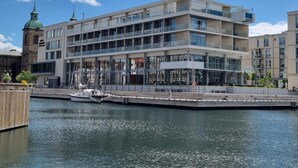 Um edifício moderno com fachada de vidro, um cais com barco e uma torre histórica ao fundo.