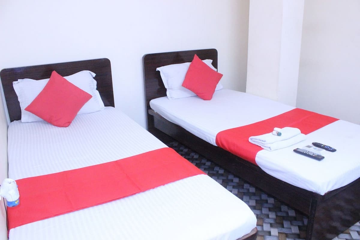 Duas camas de solteiro com almofadas vermelhas e roupa de cama branca, uma garrafa de água e dois controles remotos na cama da direita.