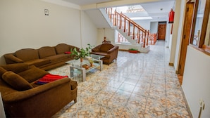 Uma sala de estar espaçosa com um sofá marrom, uma mesa de centro com objetos decorativos e piso de azulejos. Uma escada com guarda-corrimão de madeira leva a um andar superior.