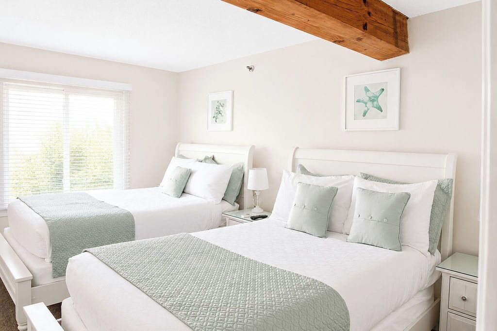 Quarto com duas camas, roupa de cama branca, viga de madeira no teto e obras de arte nas paredes.