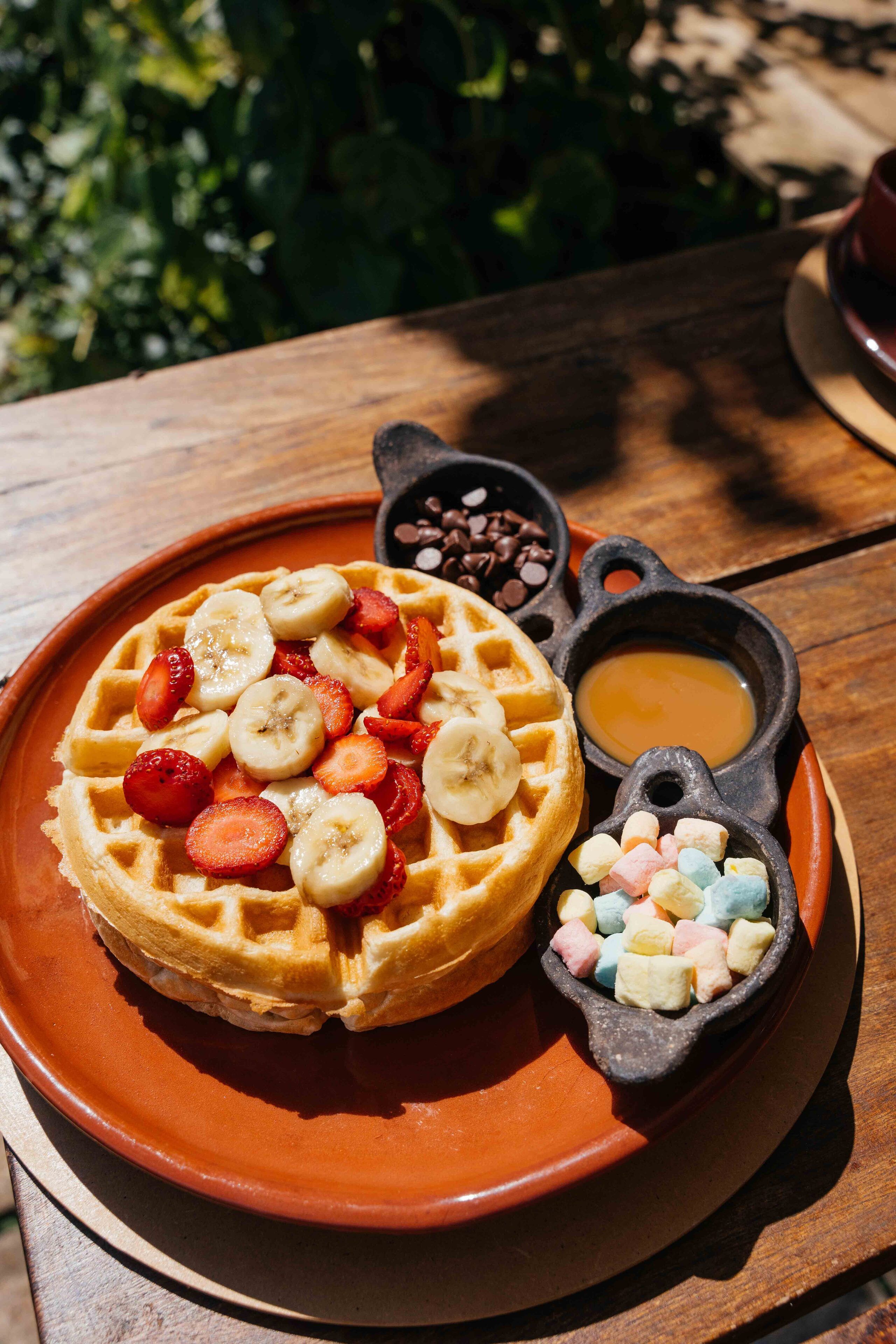 Uma pilha de waffles coberta com morangos e bananas, uma tigela pequena de marshmallows e uma xícara de calda sobre uma mesa de madeira.