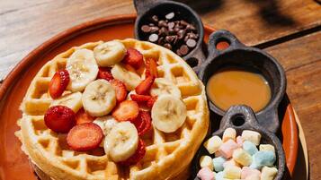 Uma pilha de waffles coberta com morangos e bananas, uma tigela pequena de marshmallows e uma xícara de calda sobre uma mesa de madeira.