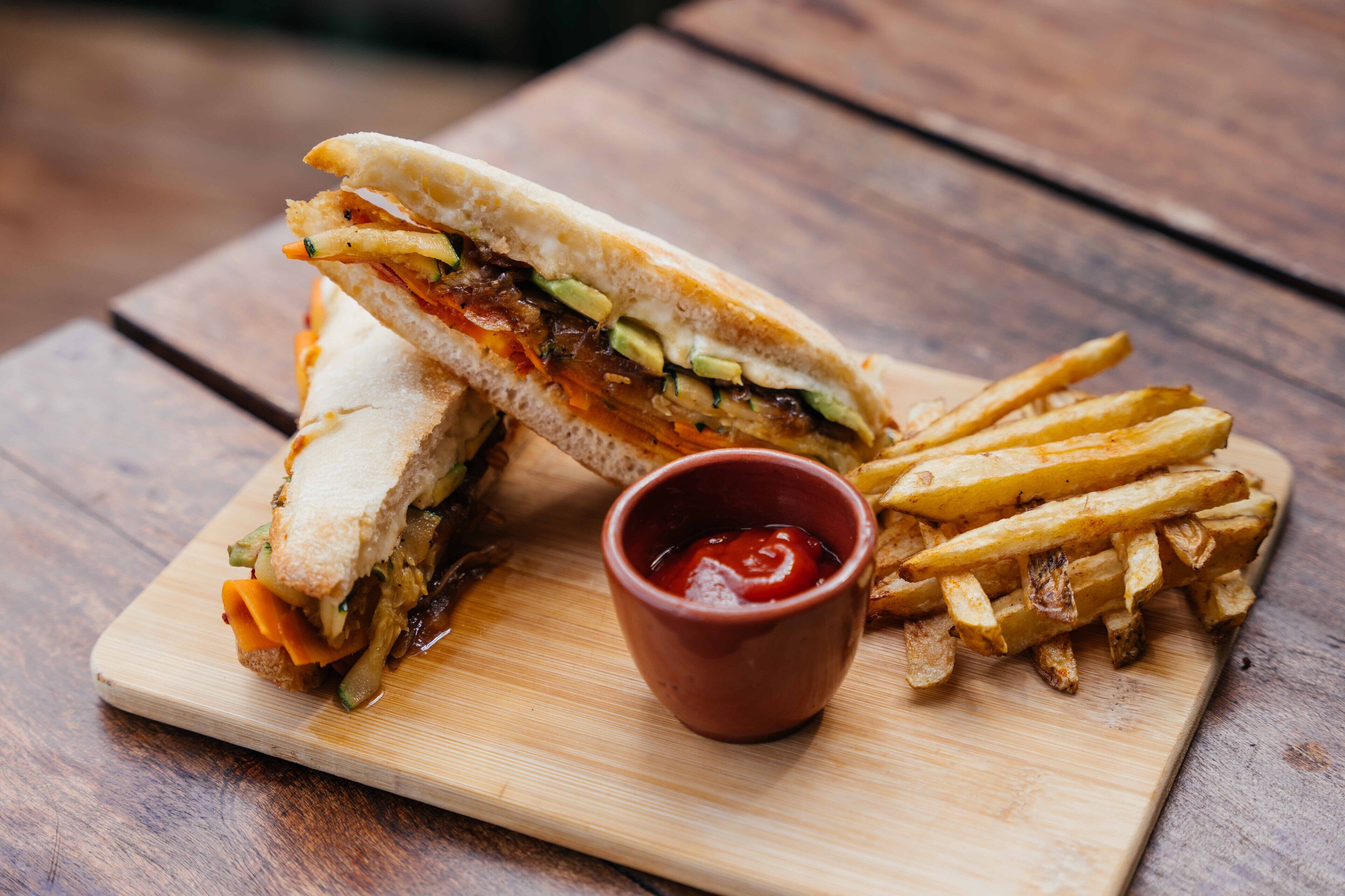 Sanduíche cortado ao meio com batatas fritas e um pequeno copo de ketchup sobre uma tábua de madeira.