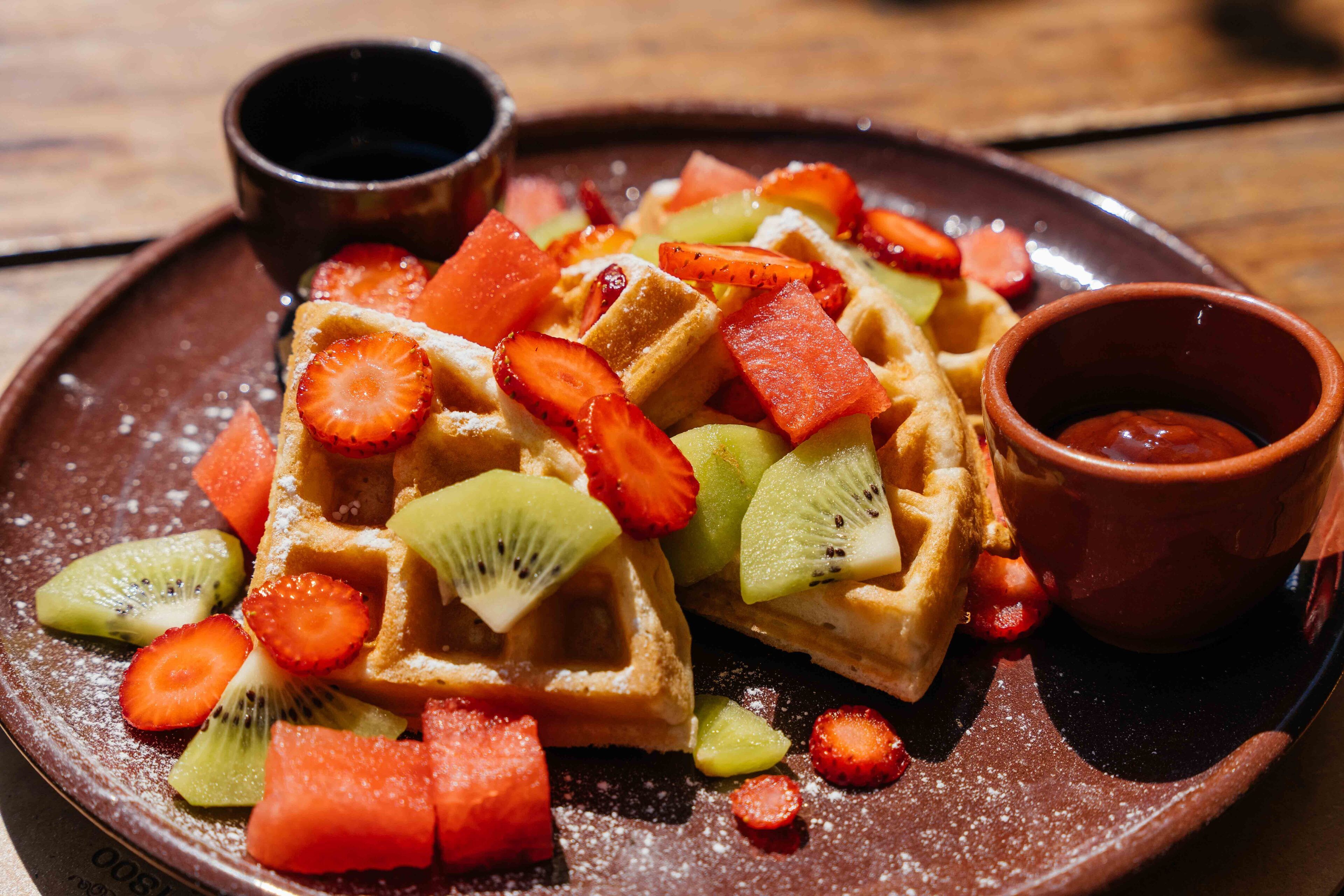 Prato de waffles coberto com morangos, kiwi e melancia, acompanhado de duas pequenas tigelas de xarope.