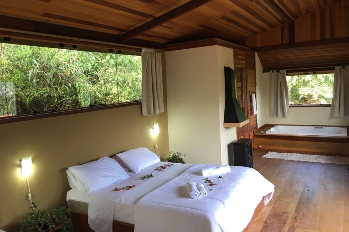 Quarto com teto de madeira, cama com roupa de cama branca, mesa de cabeceira, janela com cortinas e vista para o verde lá fora.