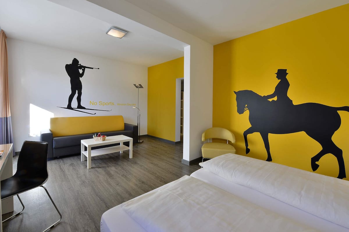Quarto de hotel com cama, sofá, mesinha, cadeira e parede com silhuetas de um esquiador e um cavaleiro.