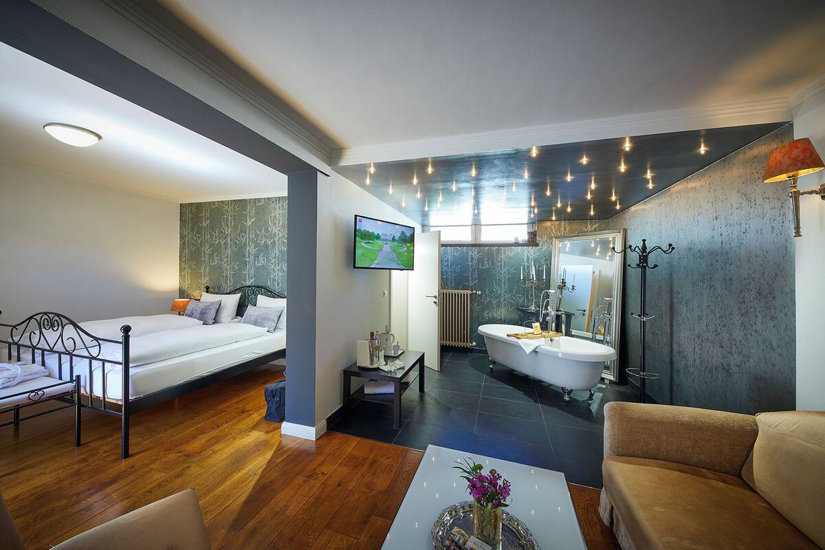 Quarto de hotel moderno com cama, sofá, mesinha com um vaso, banheiro com banheira e TV na parede.