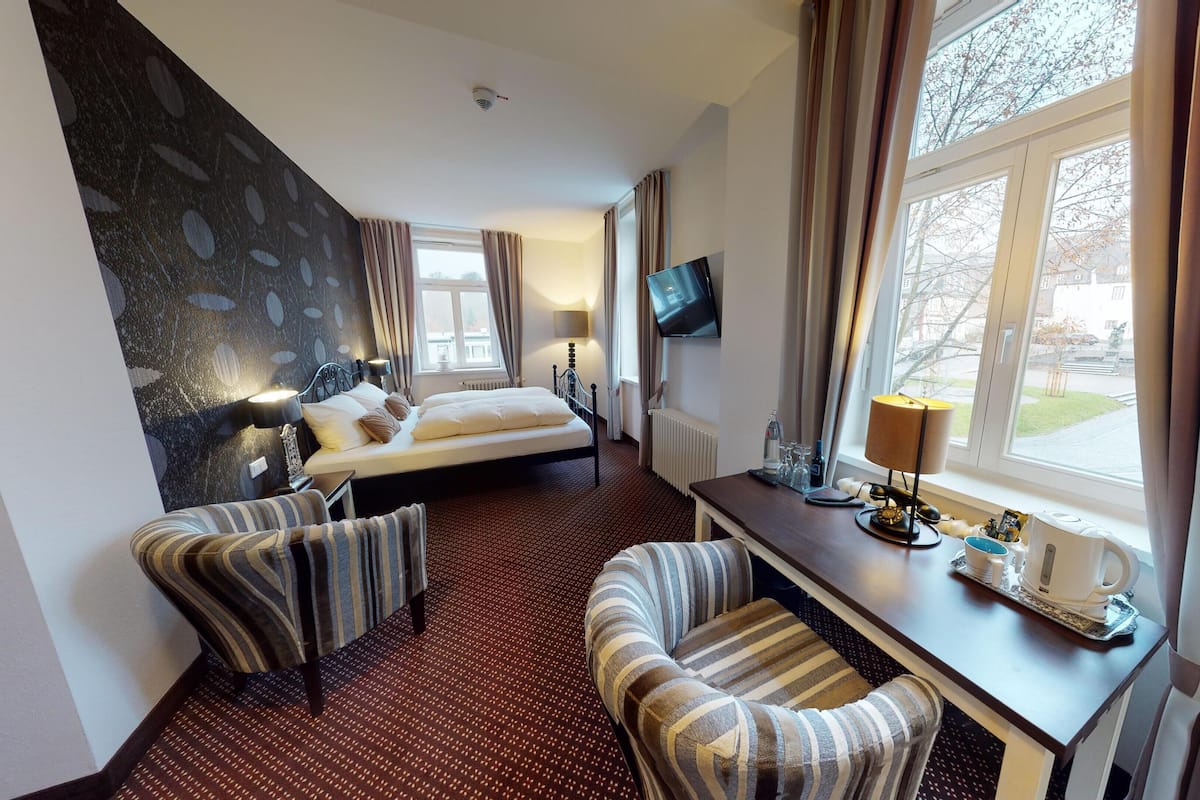 Quarto de hotel com cama, duas poltronas, escrivaninha com luminária, televisão e janela com cortinas.