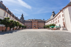 Uma praça de paralelepípedos com edifícios históricos, uma fonte central e uma mesa com guarda-sol verde.