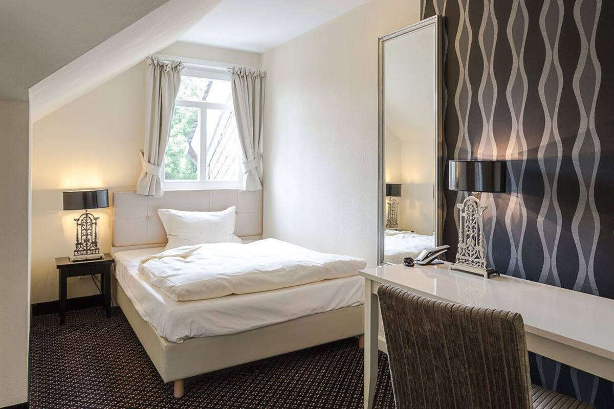 Quarto de hotel com cama, mesa, cadeira, espelho grande e janela com cortinas.