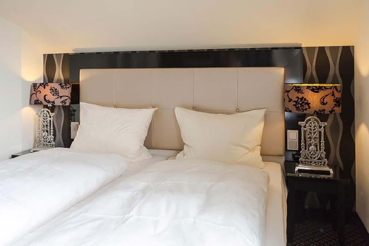 Cama bem arrumada com roupa de cama branca, cabeceira moderna e duas luminárias de parede com cúpulas estampadas.
