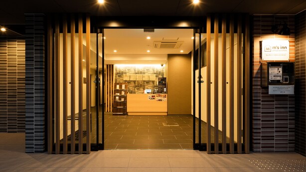 Entrada de hotel com balcão de recepção, uma placa e escadas que levam ao andar superior.