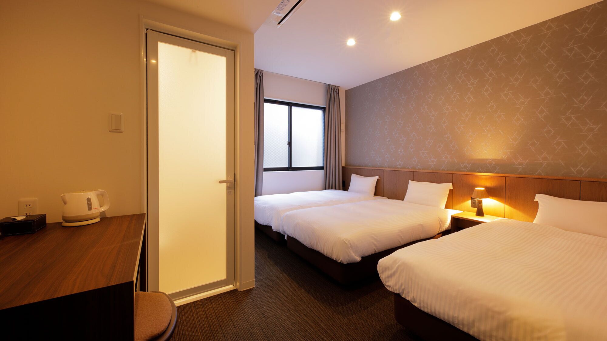 Quarto de hotel com duas camas, uma escrivaninha com chaleira, janela com cortinas e parede estampada.