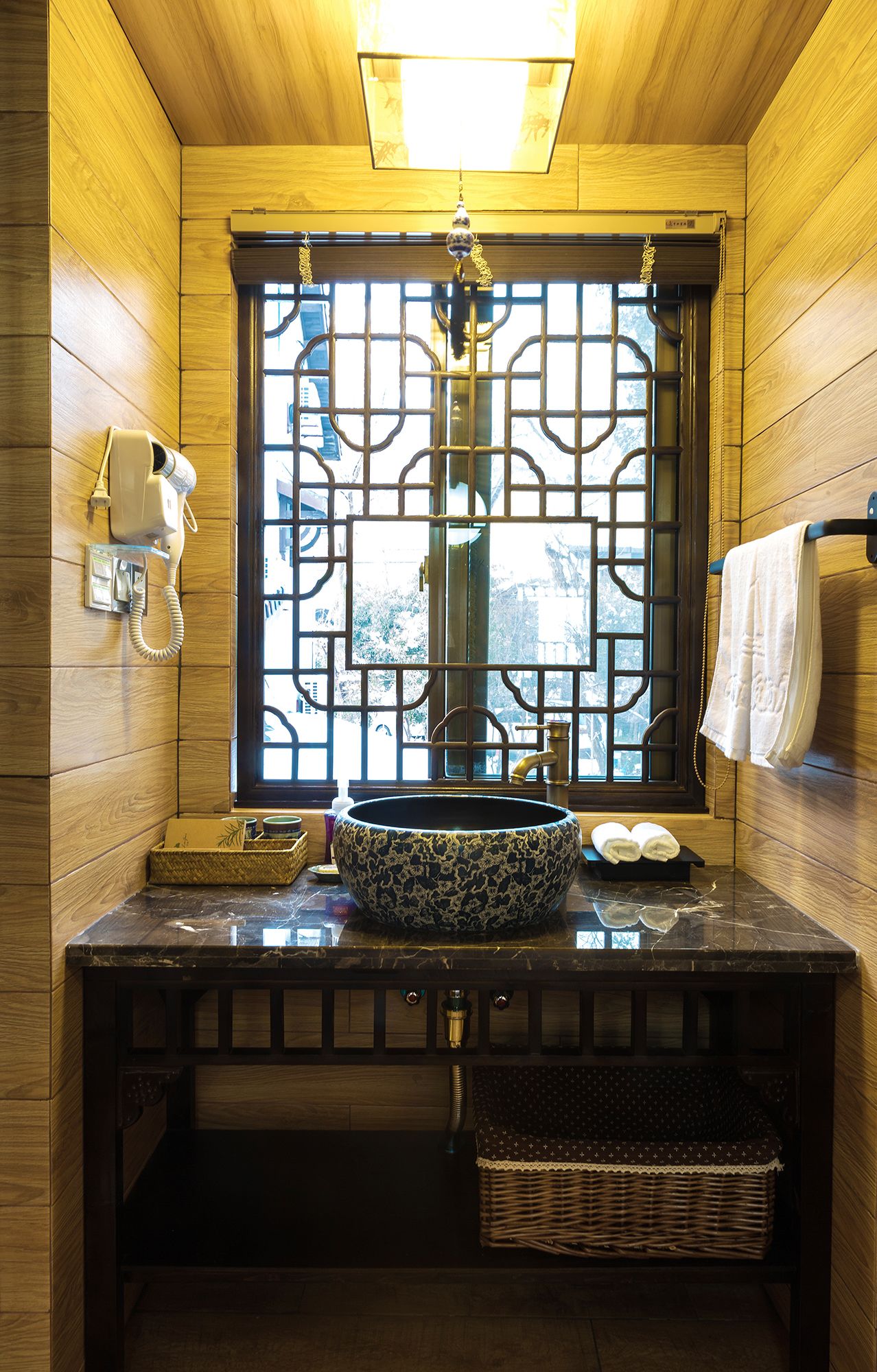 Um banheiro com decoração em madeira, uma janela ampla com grades metálicas decorativas, uma pia com uma bacia ornamental, um secador de cabelo e um porta-toalhas.