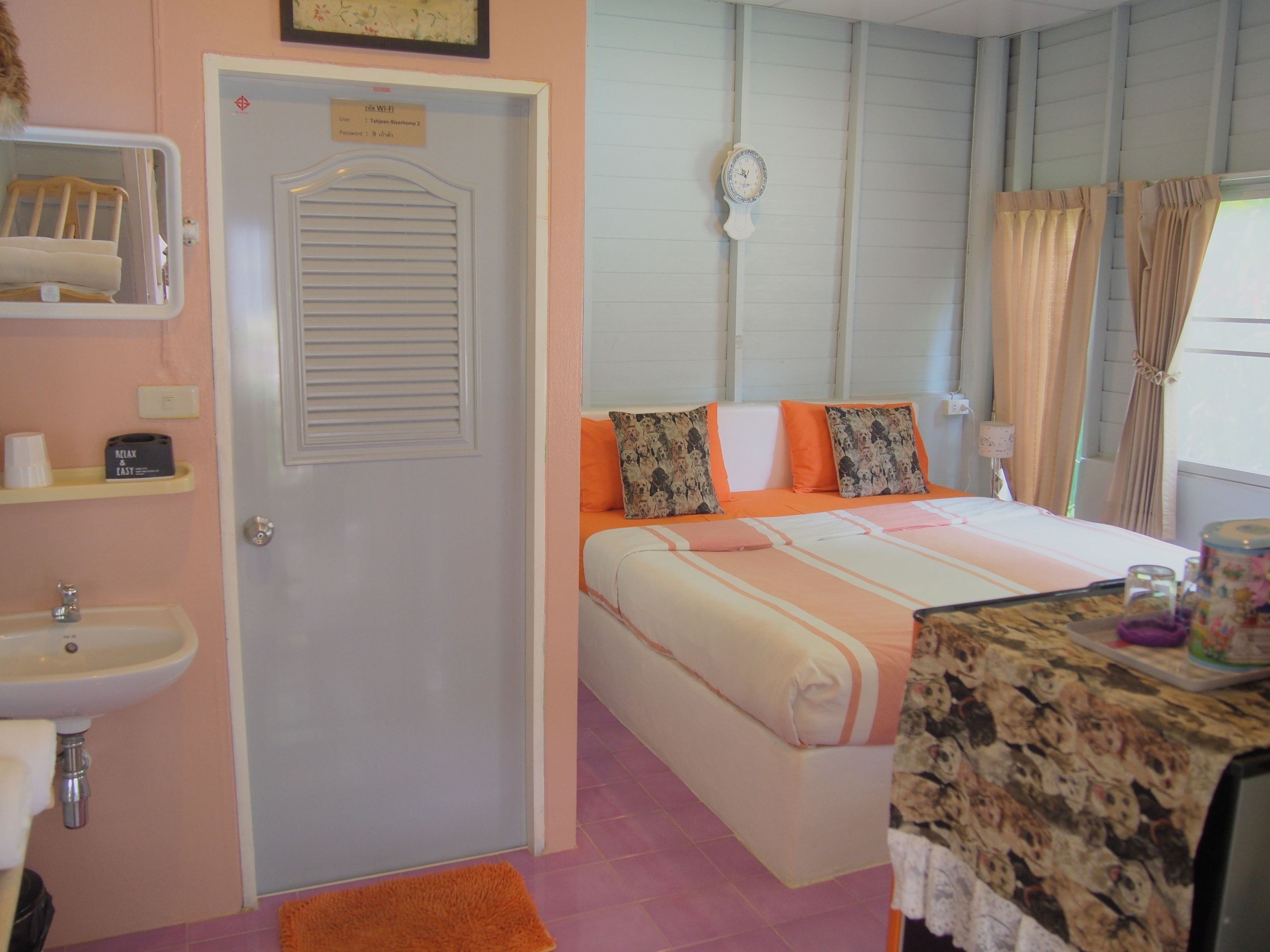 Quarto com cama, mesa de cabeceira, relógio na parede, porta e banheiro com pia e espelho.