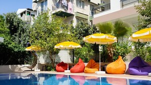 Piscina com bean bags coloridos e guarda-sóis, cercada por vegetação e um edifício de vários andares ao fundo.