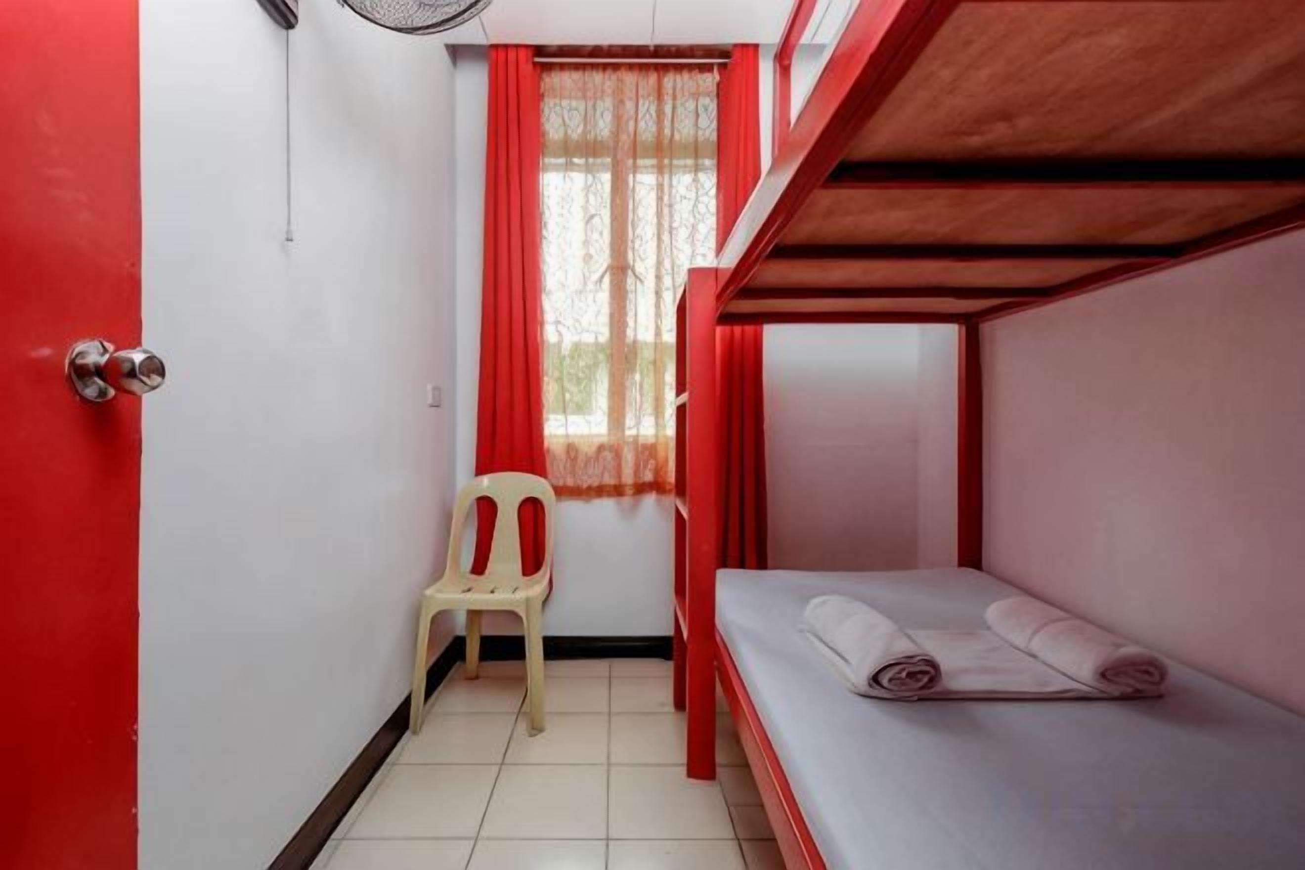 Um quarto pequeno com uma cama de solteiro, uma cadeira e uma janela com cortinas vermelhas.