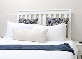 Uma cama com almofadas brancas e estampadas, um cobertor azul escuro e uma cabeceira branca.