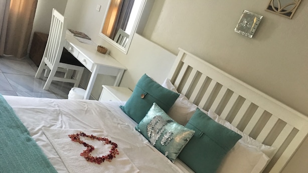 Cama bem arrumada com um coração decorativo no travesseiro, uma pequena escrivaninha com cadeira e uma mesa de cabeceira com telefone.