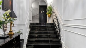 Um chão xadrez em preto e branco, uma escada com corrimão preto, um candelabro, um vaso de planta e um quadro emoldurado na parede.